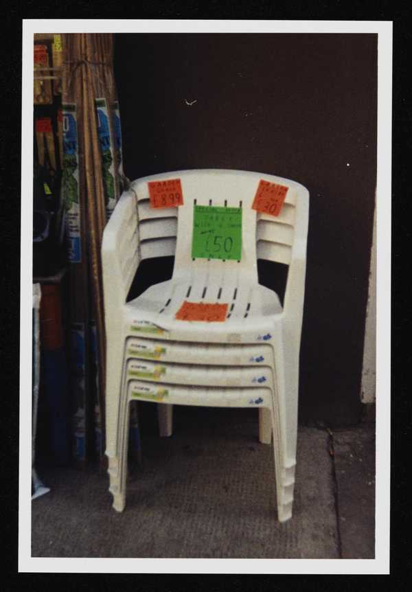 Colour photograph of white plastic garden chairs stacked up outside a ...